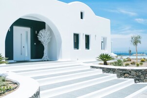 Um edifício moderno branco com entrada curva, um caminho de pedra que leva à porta e vista para o mar ao fundo.