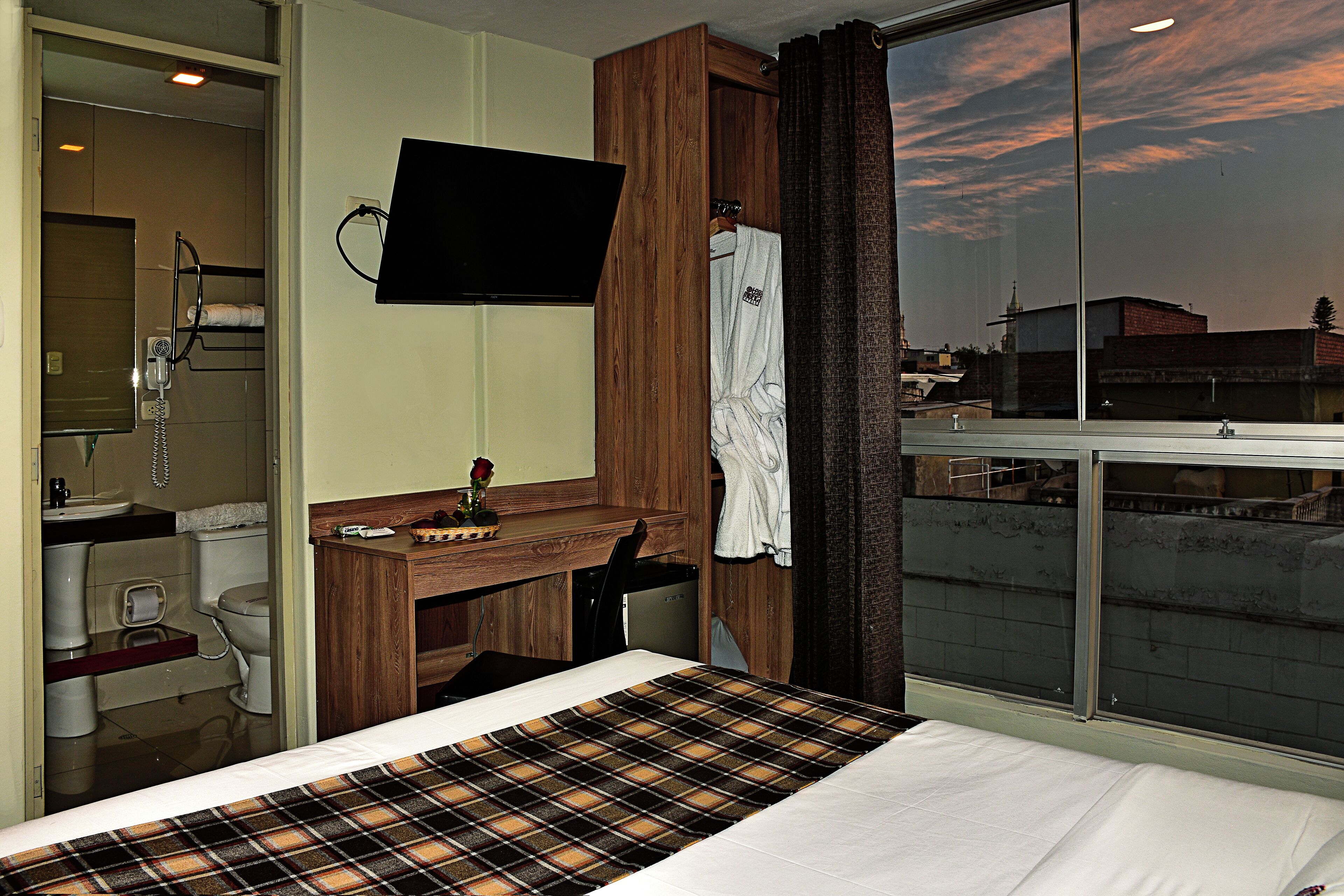 Quarto de hotel com cama, TV fixada na parede, secretária de madeira e vista para a cidade através de amplas janelas.