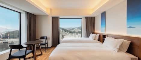 Quarto de hotel com duas camas, uma mesa pequena e uma cadeira. Uma janela grande oferece vista para a cidade.