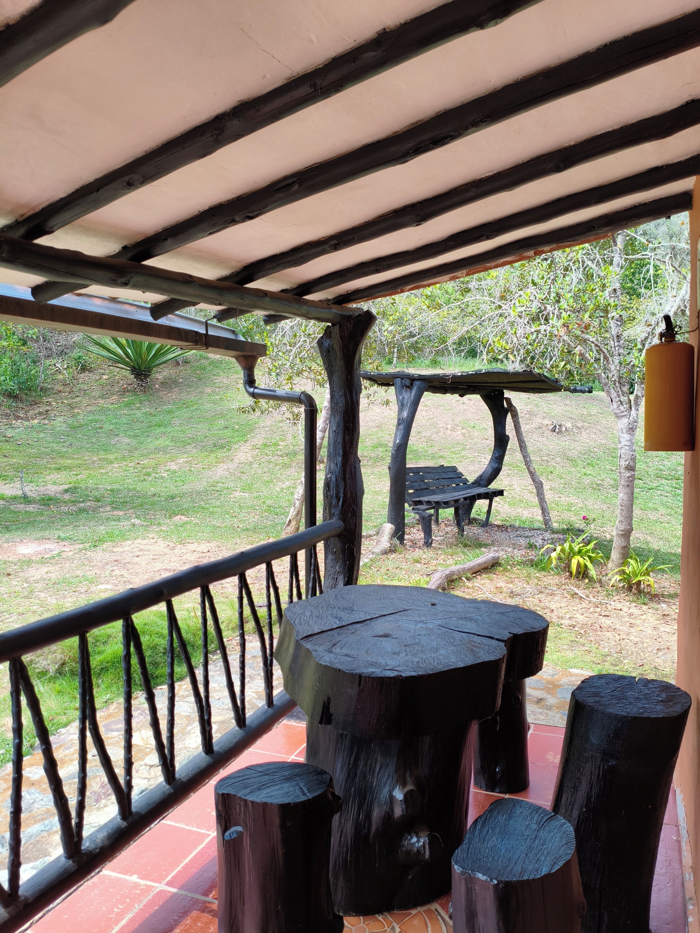 Área de estar coberta ao ar livre, com mesa e cadeiras, delimitada por um cercado de madeira e vista para um gramado com árvores.