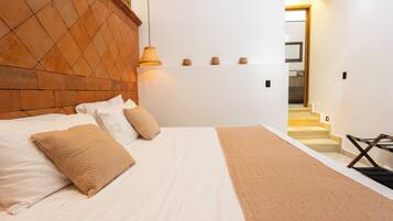 Quarto com cama, almofadas, luminária de cabeceira, cabeceira de madeira, prateleira pequena com objetos decorativos e escada que leva a um banheiro.