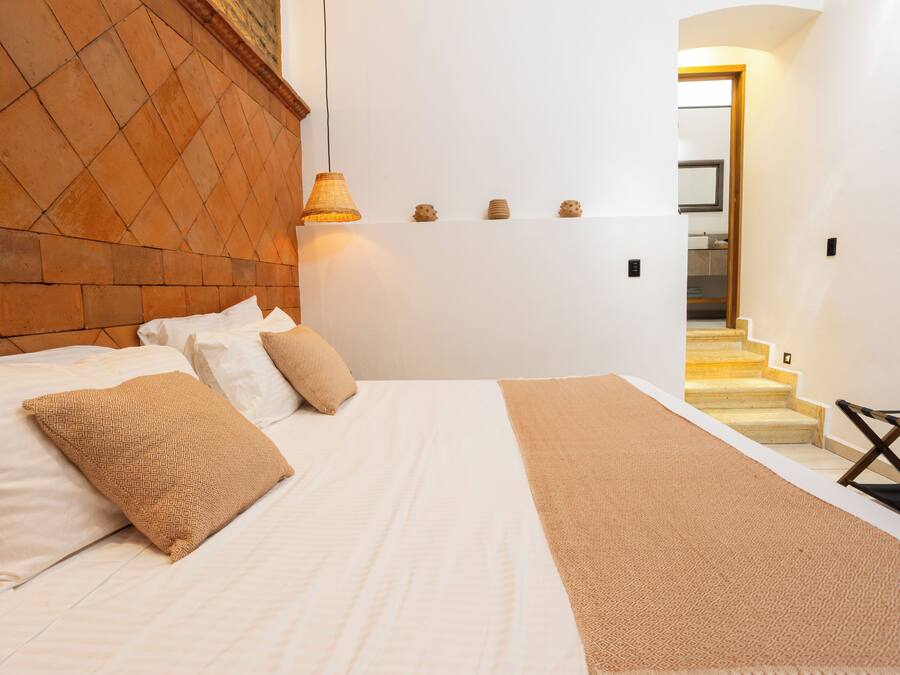 Quarto com cama, almofadas, luminária de cabeceira, cabeceira de madeira, prateleira pequena com objetos decorativos e escada que leva a um banheiro.
