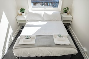 Cama bem arrumada com lençóis e toalhas brancos, um cobertor cinza e uma janela com vista para prédios.