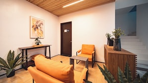 Uma área de espera moderna com teto de madeira, um quadro abstrato emoldurado, uma mesa consola de madeira com um vaso e uma área de estar com cadeiras laranja.