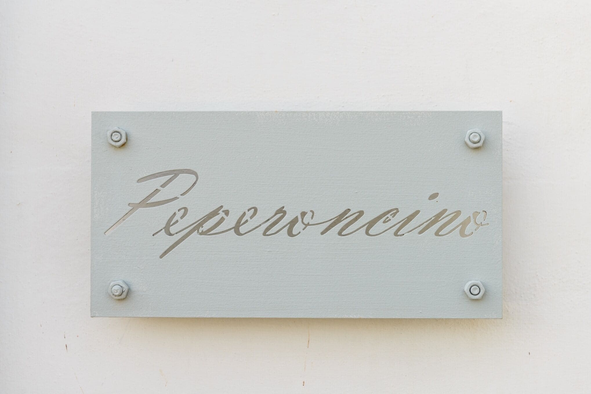Uma placa cinza clara com a palavra "Peppermint" em letra cursiva, fixada na parede com quatro parafusos visíveis.