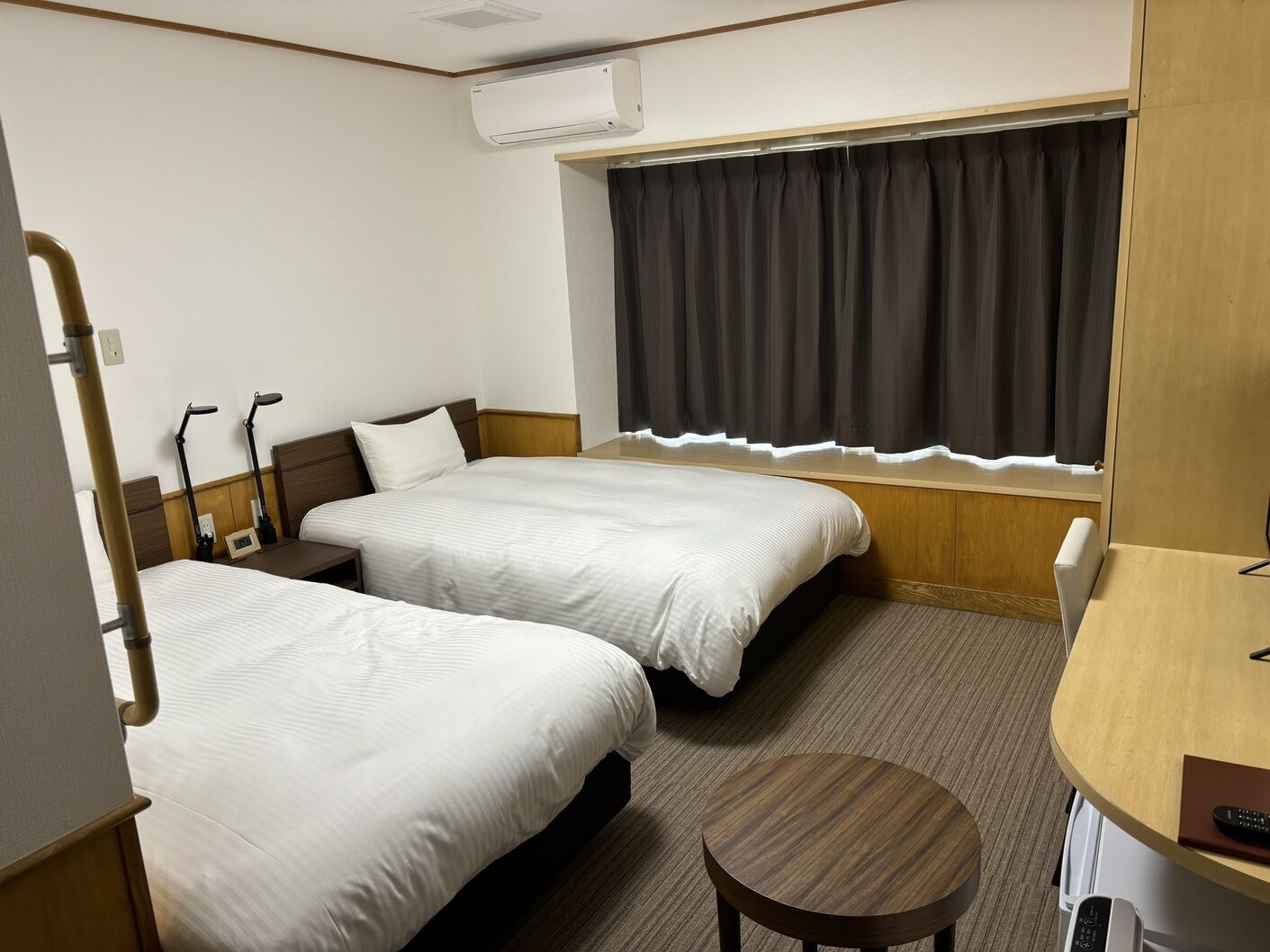 Quarto de hotel com duas camas, uma escrivaninha, uma cadeira, ar condicionado e uma janela com cortinas.
