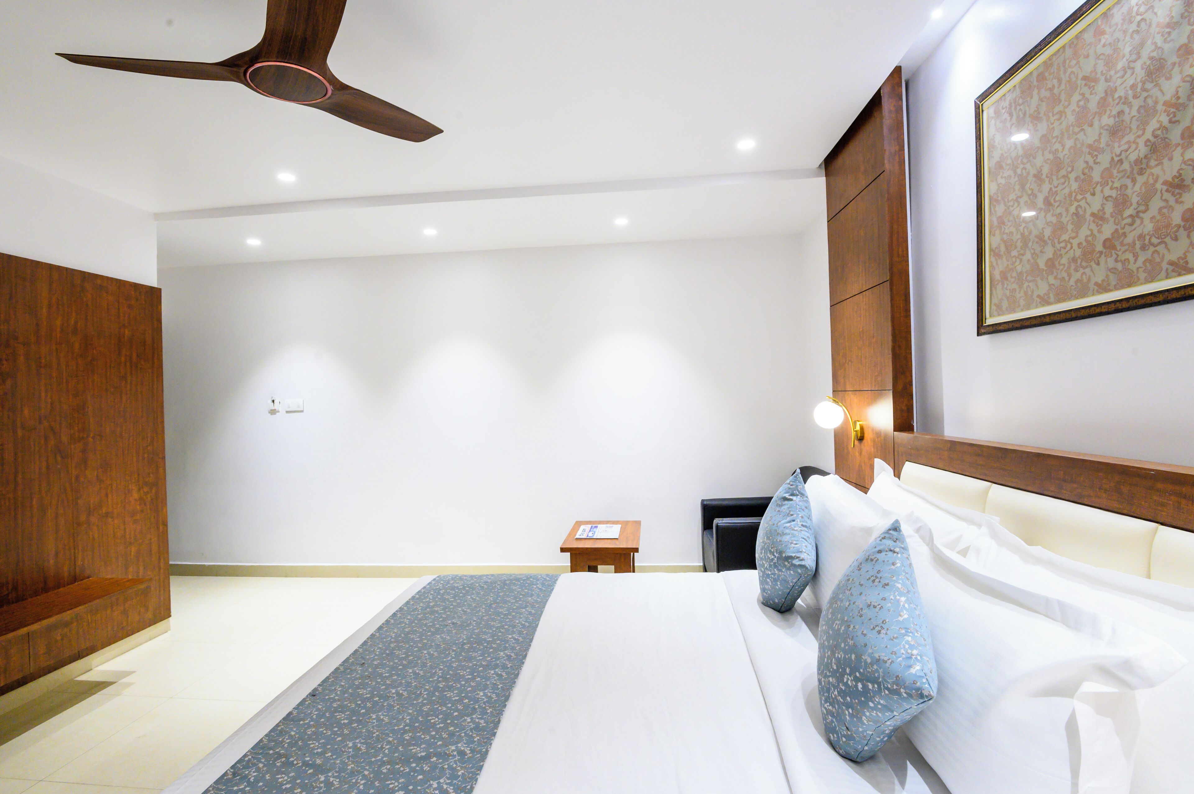 Quarto de hotel com uma cama grande, ventilador de teto, uma mesinha e luminária na parede.