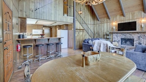 Uma sala de estar ampla com uma grande mesa de jantar de madeira, uma lareira, um espaço bar com banquetas e uma escadaria para um andar superior.