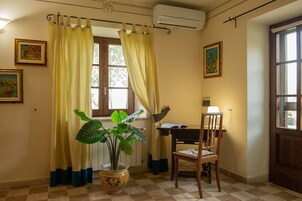 Um quarto com chão xadrez, um vaso de planta, uma cadeira, uma escrivaninha com abajur e duas janelas com cortinas.