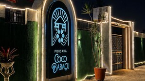 Vista noturna da entrada de um prédio com um letreiro de neon que diz 'POUSADA Coco BABAÇU' e uma planta em vaso.