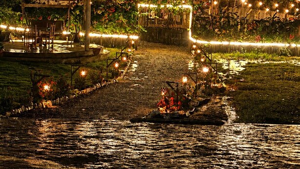 Ambiente externo noturno com um espelho d'água, estruturas iluminadas e um gazebo.