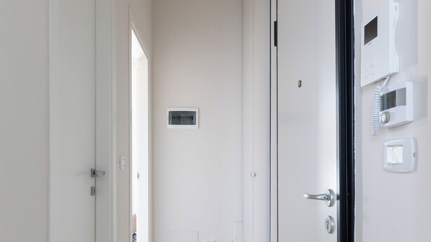 Um corredor com piso de madeira, paredes brancas e uma porta com moldura preta à direita.