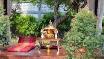 Um altar tailandês tradicional com uma estátua dourada, um cálice e flores, colocado sobre uma mesa de madeira num jardim exuberante.