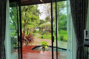 Vista através de uma porta de vidro para um jardim com lago, plantas em vasos e um caminho.