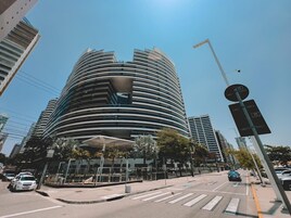 Um edifício moderno de vários andares com fachada curva, cercado por palmeiras e outras instalações urbanas.