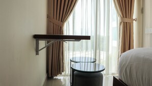 Quarto de hotel com cama, uma mesinha redonda, prateleira fixada na parede e cortinas leves.