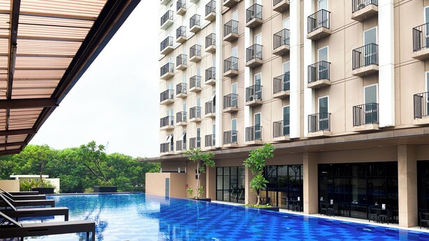 Piscina com piso azulejado e espreguiçadeiras, cercada por um edifício com varandas.