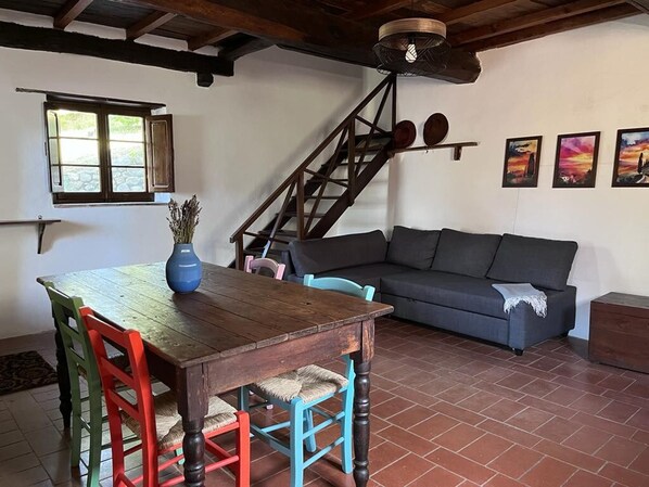Uma área de refeições com mesa de madeira e cadeiras coloridas, um sofá cinza e uma escada com ventilador.
