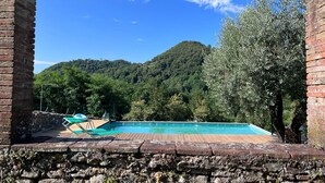 Piscina com deck de madeira, cercada por muros de pedra e oliveiras, com uma montanha ao fundo.