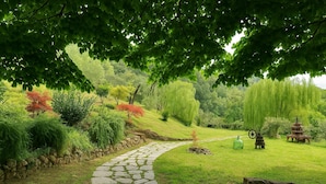 Uma trilha de pedra serpenteia por um jardim exuberante, com uma estrutura em estilo pagode e uma grande garrafa verde.
