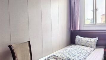 Uma cama de solteiro com colcha florida em quarto com paredes claras e janela com cortinas roxas.