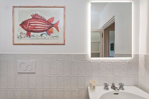 Um banheiro com uma ilustração de peixe emoldurada, um espelho, uma pia e um vaso sanitário com uma xícara em cima.