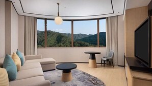 Quarto de hotel moderno com uma grande janela que oferece vista para as montanhas, um sofá, uma mesa de centro redonda e uma televisão de tela plana.