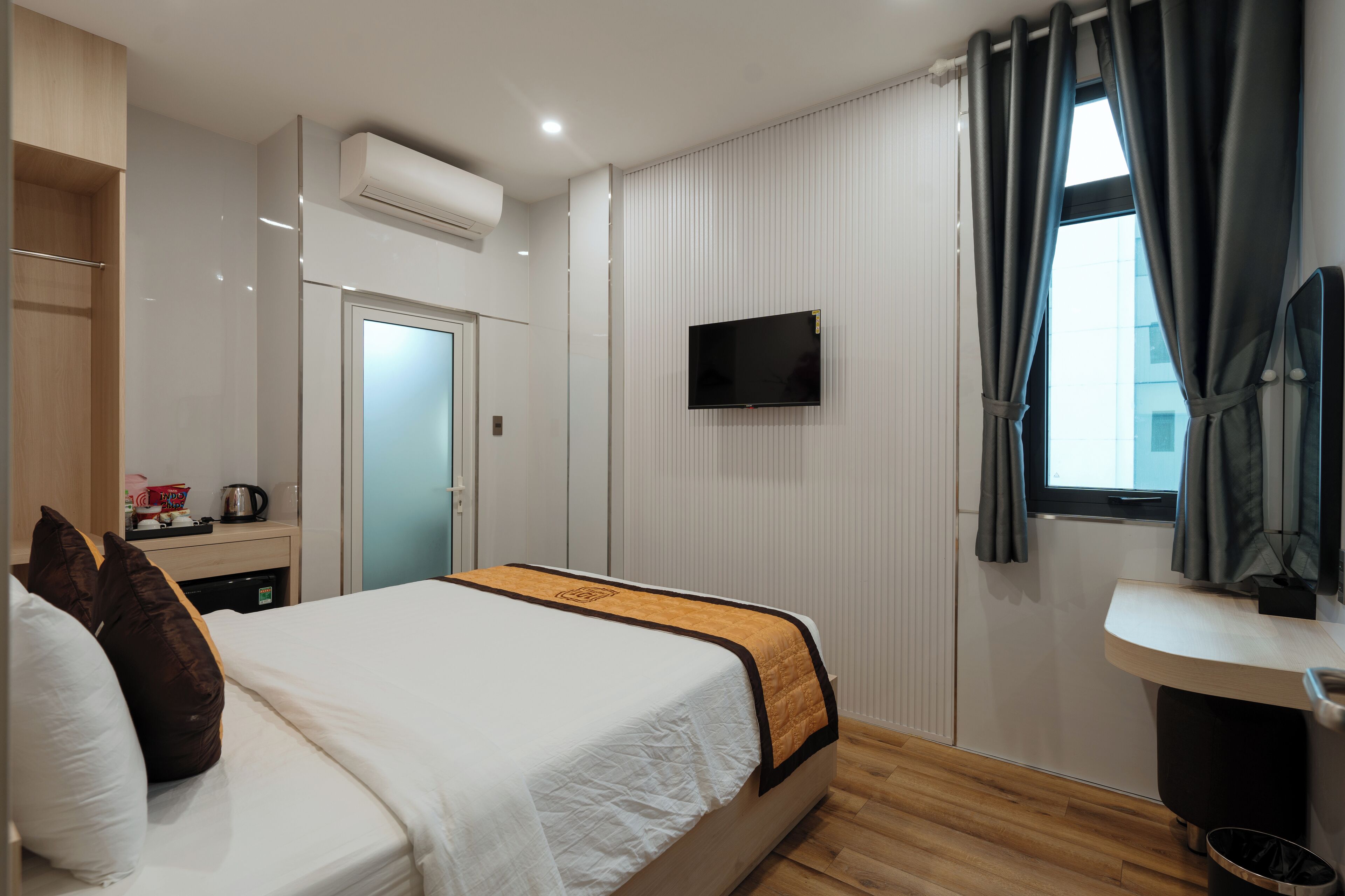 Quarto de hotel moderno com cama, televisão fixada na parede, pequena escrivaninha com cafeteira e janela com cortinas.