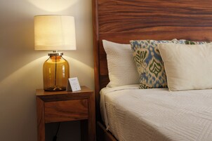 Cama bem arrumada com colcha branca e almofadas estampadas, mesa de cabeceira de madeira com abajur e cabeceira de madeira.