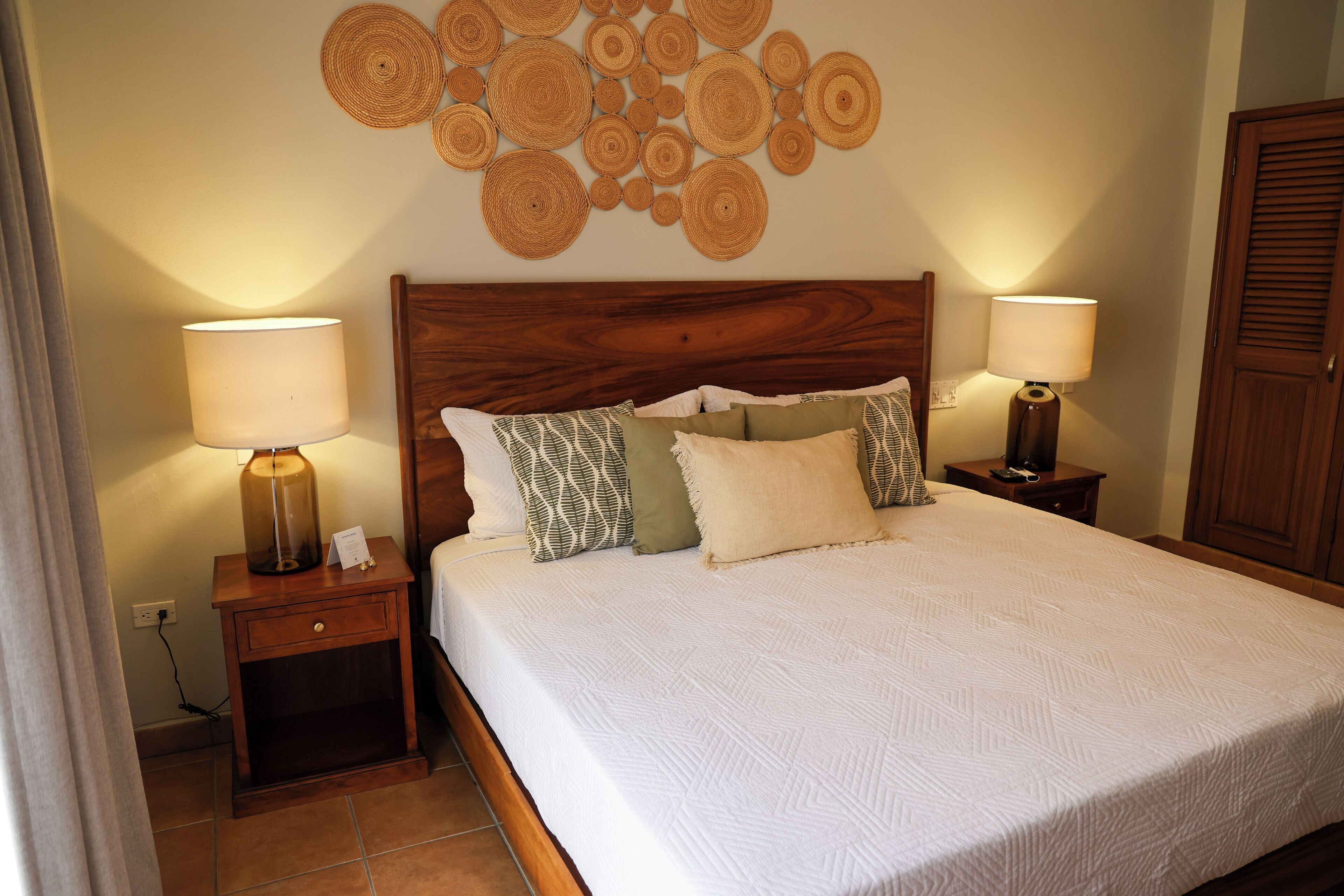 Uma cama bem arrumada com colcha branca, duas mesas de cabeceira com abajures, um painel de cabeceira de madeira e um adorno na parede.