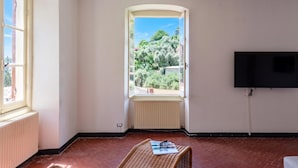 Um quarto com uma janela grande, um radiador, uma televisão e uma cadeira de vime.