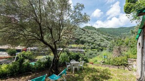 Paisagem rural com uma grande árvore, uma mesa e cadeiras brancas e um muro de pedra.