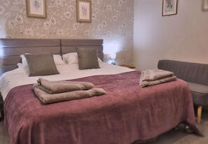Uma cama bem arrumada com colcha roxa e duas almofadas, um sofá cinza e papel de parede floral.