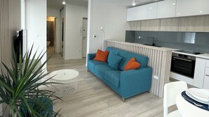 Uma sala de estar moderna com um sofá azul, uma mesa de jantar e uma área de cozinha com armários brancos e forno embutido.