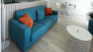Uma sala de estar moderna com um sofá azul, uma área de jantar com cadeiras brancas e uma cozinha com armários brancos e uma luminária pendente.