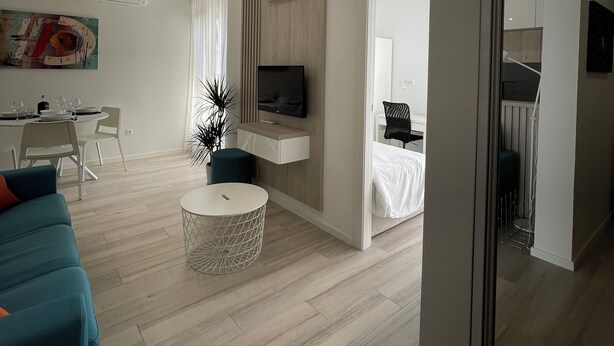 Uma sala de estar moderna com televisão de tela plana, uma área de jantar com mesa e cadeiras, e um quarto com cama e mesa de cabeceira.