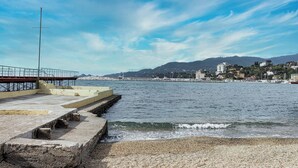 Uma orla costeira com calçada de concreto, grades de metal e uma praia de pedras.