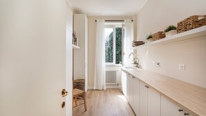 Um corredor estreito com piso de madeira, paredes brancas e uma janela com cortinas brancas. Há uma cadeira de vime e uma prateleira com cestos e plantas.