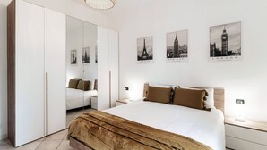 Quarto com cama grande, guarda-roupa e mesa de cabeceira. Há quadros pendurados na parede com pontos turísticos famosos.