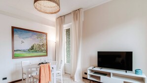 Uma sala de estar moderna com uma televisão de tela plana, mobiliário branco, uma pintura de paisagem e uma janela grande com cortinas.