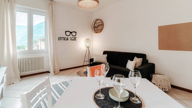 Uma sala de estar moderna com uma mesa branca posta para refeições, um sofá preto e uma janela grande com cortinas.