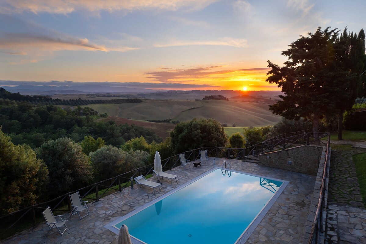Piscina com espreguiçadeiras e um deck de pedra, cercada por vegetação e uma vista panorâmica de colinas e um pôr do sol.