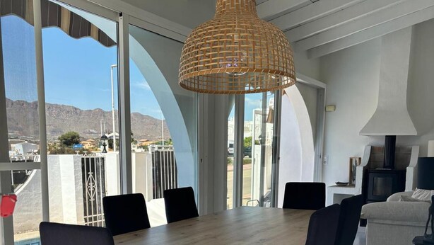 Uma sala de jantar com mesa de madeira e cadeiras pretas, janelas amplas com vista para o mar e um candeeiro de vime pendente.