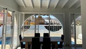 Um quarto espaçoso com uma mesa grande e cadeiras, um ventilador de teto e janelas amplas com vista para uma piscina e edifícios.