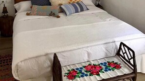 Cama bem arrumada com edredom branco e várias almofadas, cabeceira de madeira e uma cadeira com toalha de mesa florida.