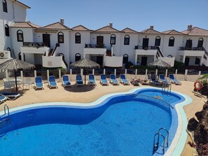 Piscina com espreguiçadeiras e guarda-sóis, cercada por um edifício com várias varandas e um telhado ladrilhado.