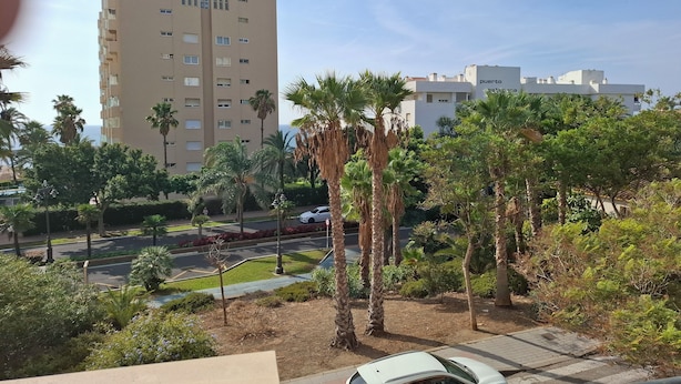Zona residencial com um prédio alto cor de bege, palmeiras e um carro branco estacionado na rua.