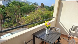 Varanda com mesa e cadeiras, vaso com flores e vista para áreas verdes e edifícios.
