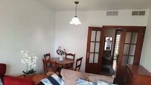 Sala de estar com um sofá bege, poltrona vermelha e mesa de jantar de madeira com cadeiras. Há um vaso com flores sobre a mesa e um lustre no teto.