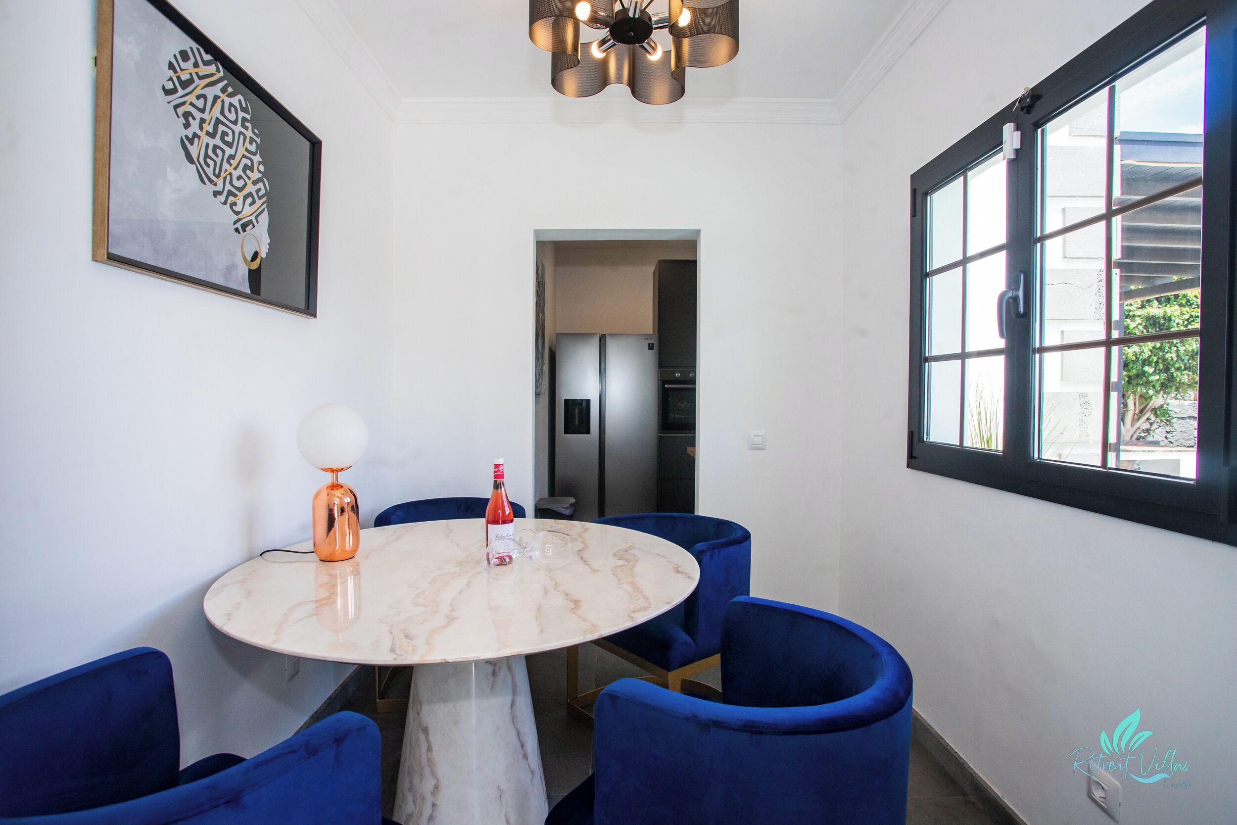 Uma sala de jantar com uma mesa redonda de mármore, cadeiras azuis, um candelabro e uma obra de arte emoldurada na parede.